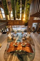 Um lobby espaçoso com uma grande mesa de madeira rodeada por cadeiras, uma área de estar com sofás e vista para o exterior através de grandes janelas.