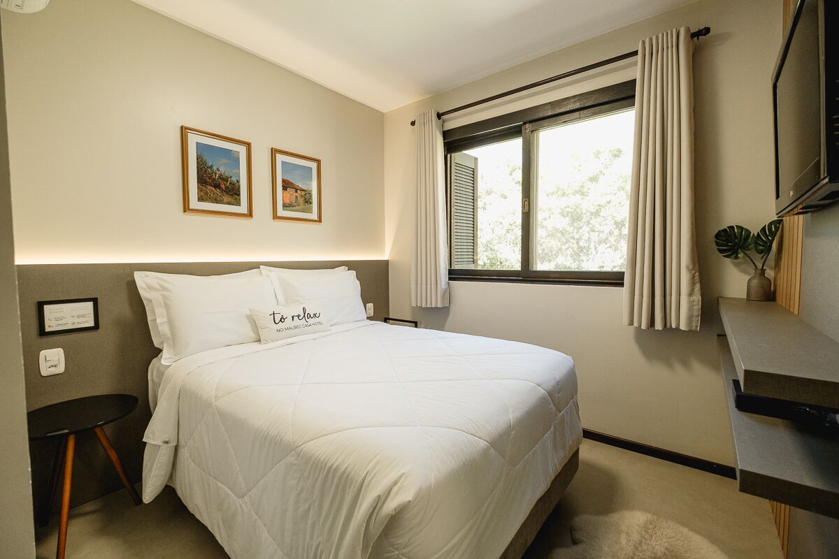 Quarto de hotel com uma cama grande, uma pequena mesa de escritório, uma janela com cortinas e duas fotos emolduradas na parede.