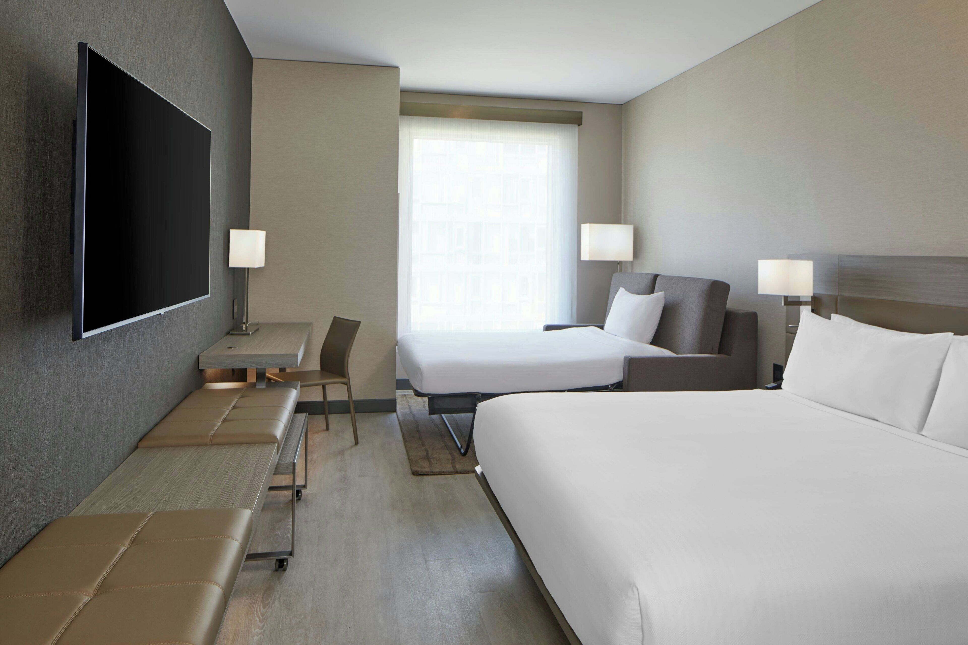 Quarto de hotel moderno com uma cama grande, uma mesa com cadeira, uma TV de tela plana e duas luminárias de cabeceira.