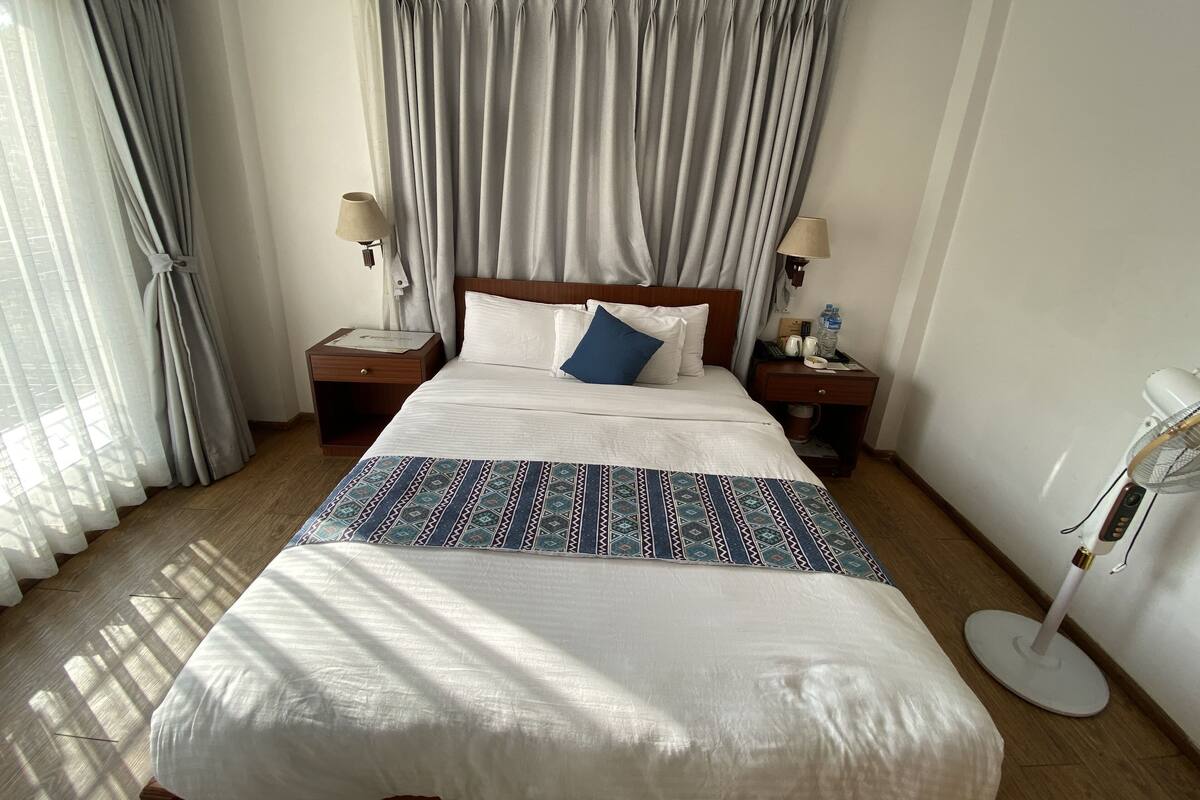Quarto de hotel com uma cama, duas mesas de cabeceira, uma luminária, um ventilador e uma janela com cortinas.