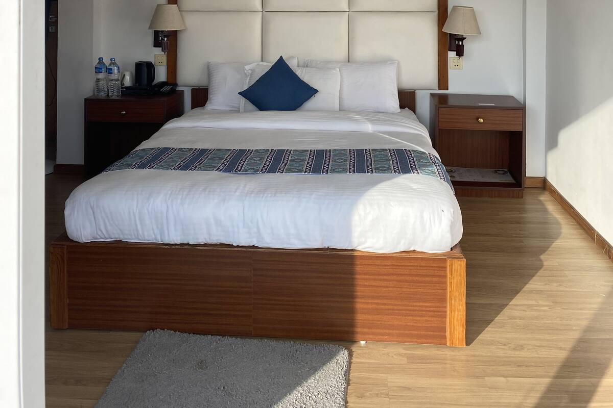 Quarto de hotel bem arrumado com uma cama grande, cabeceira de madeira, duas mesas de cabeceira com luminárias e uma pequena mesa de cabeceira com uma lâmpada.