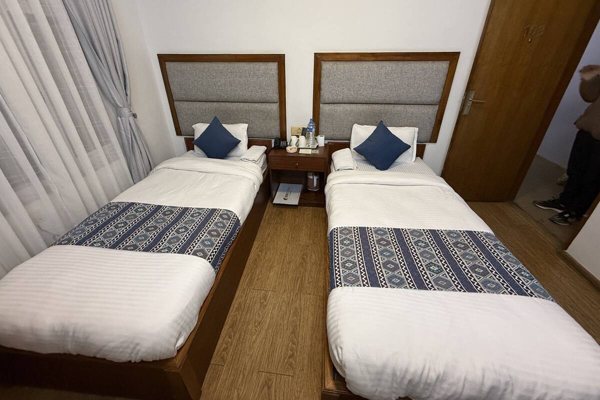 Quarto de hotel com duas camas de solteiro, mesa de cabeceira com um telefone e uma garrafa, e guarda-roupa de madeira.