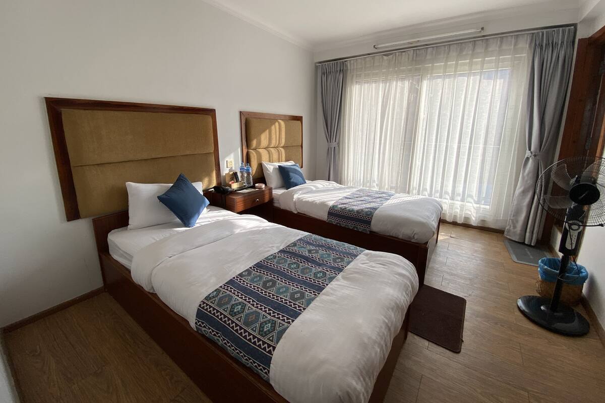 Quarto de hotel com duas camas, uma mesa de cabeceira com abajur, um ventilador e uma janela com cortinas.