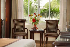 Quarto de hotel com duas cadeiras xadrez, uma mesa pequena com um vaso de flores e vista para palmeiras através de grandes janelas.