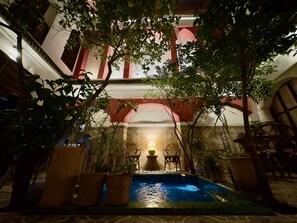 Uma área interna com piscina, chão revestido de azulejos, rodeada por plantas em vasos e mobiliário de madeira.