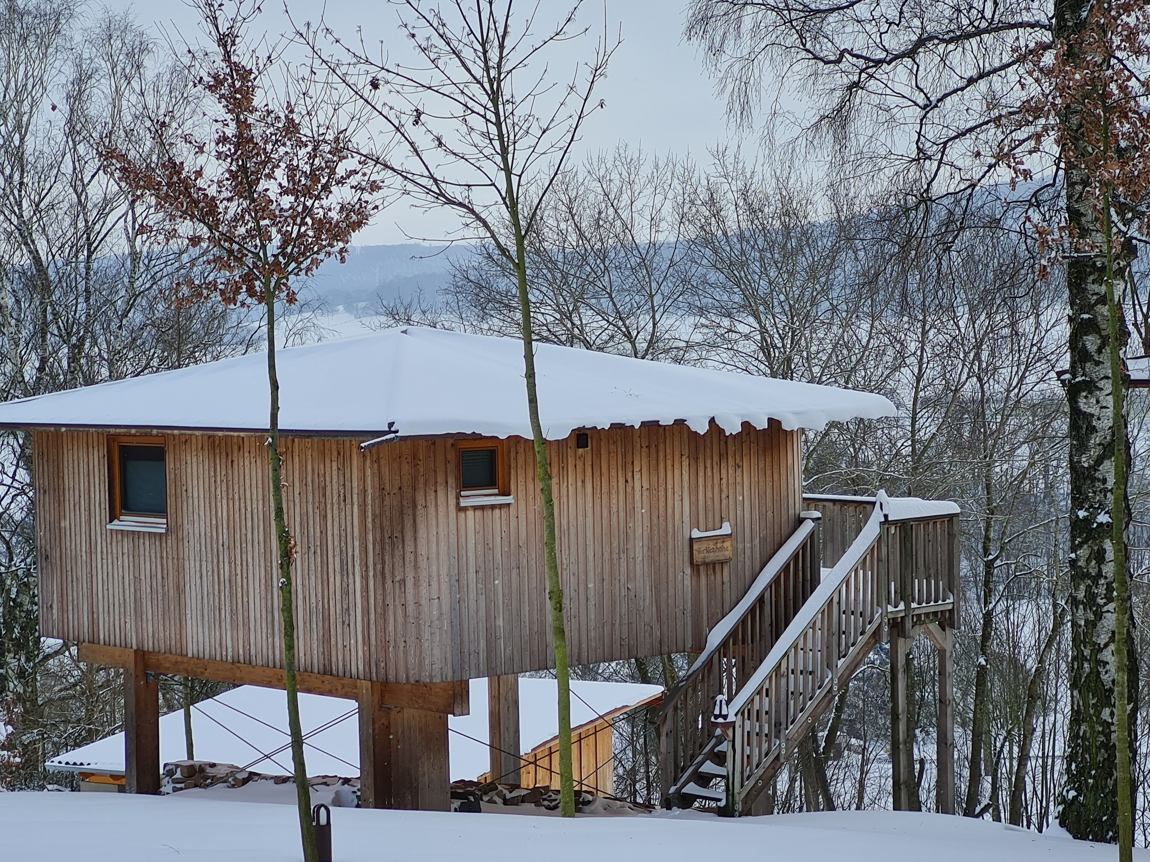 Uma cabana de madeira com telhado coberto de neve, uma pequena janela e uma escadaria que leva até ela.
