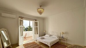 Quarto com cama, duas mesas de cabeceira, um candelabro, um espelho, uma cadeira e vista para o exterior.