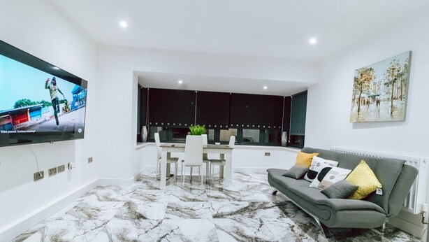 Uma sala de estar moderna com piso de mármore, um sofá cinza com almofadas, uma televisão de tela plana e uma área de jantar com uma mesa e cadeiras brancas.