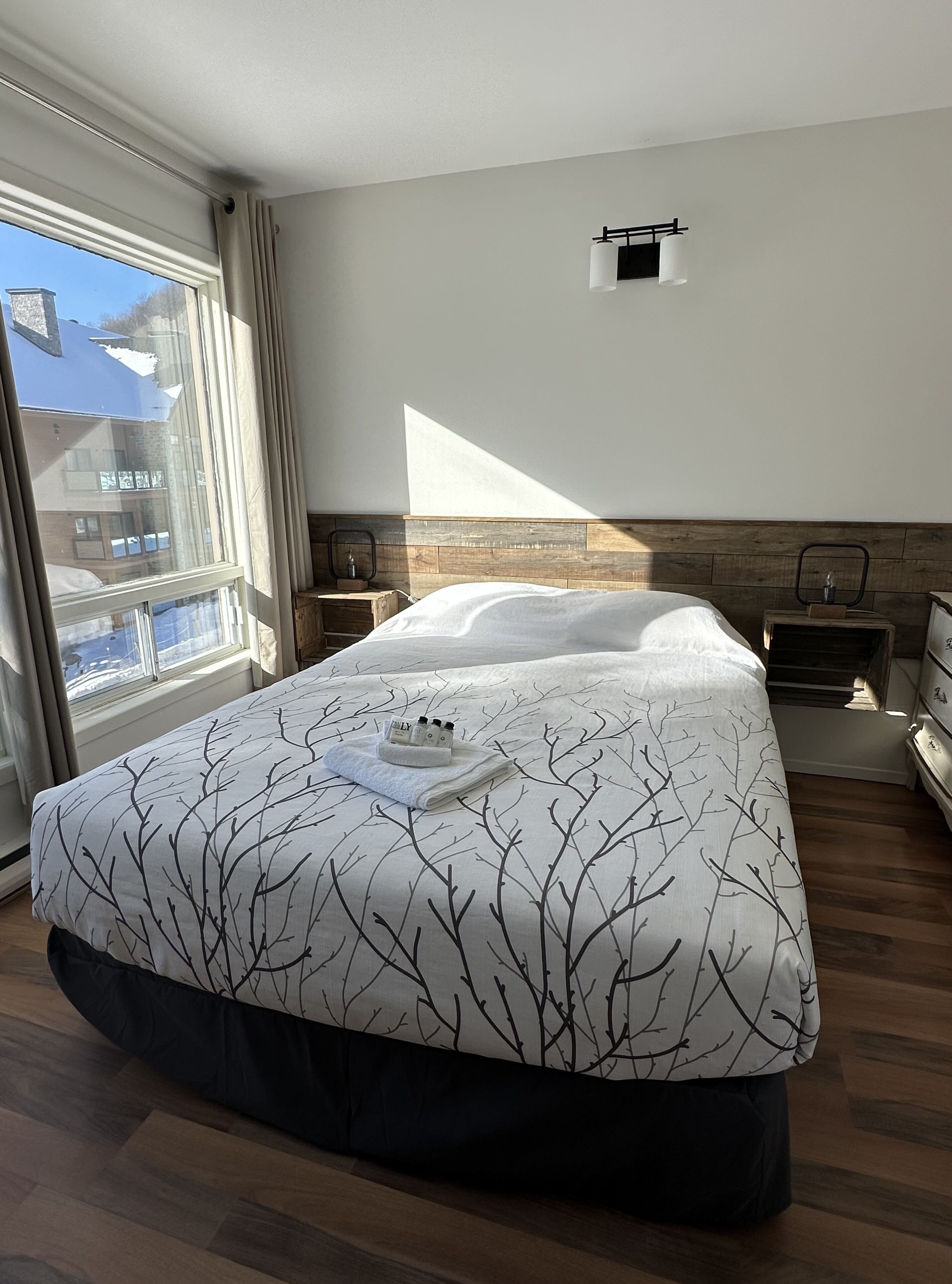 Quarto de hotel moderno com uma cama grande, cabeceira de madeira, uma mesa de cabeceira com abajur, uma pequena escrivaninha com cadeira e uma janela ampla que oferece vista para edifícios e céu azul.