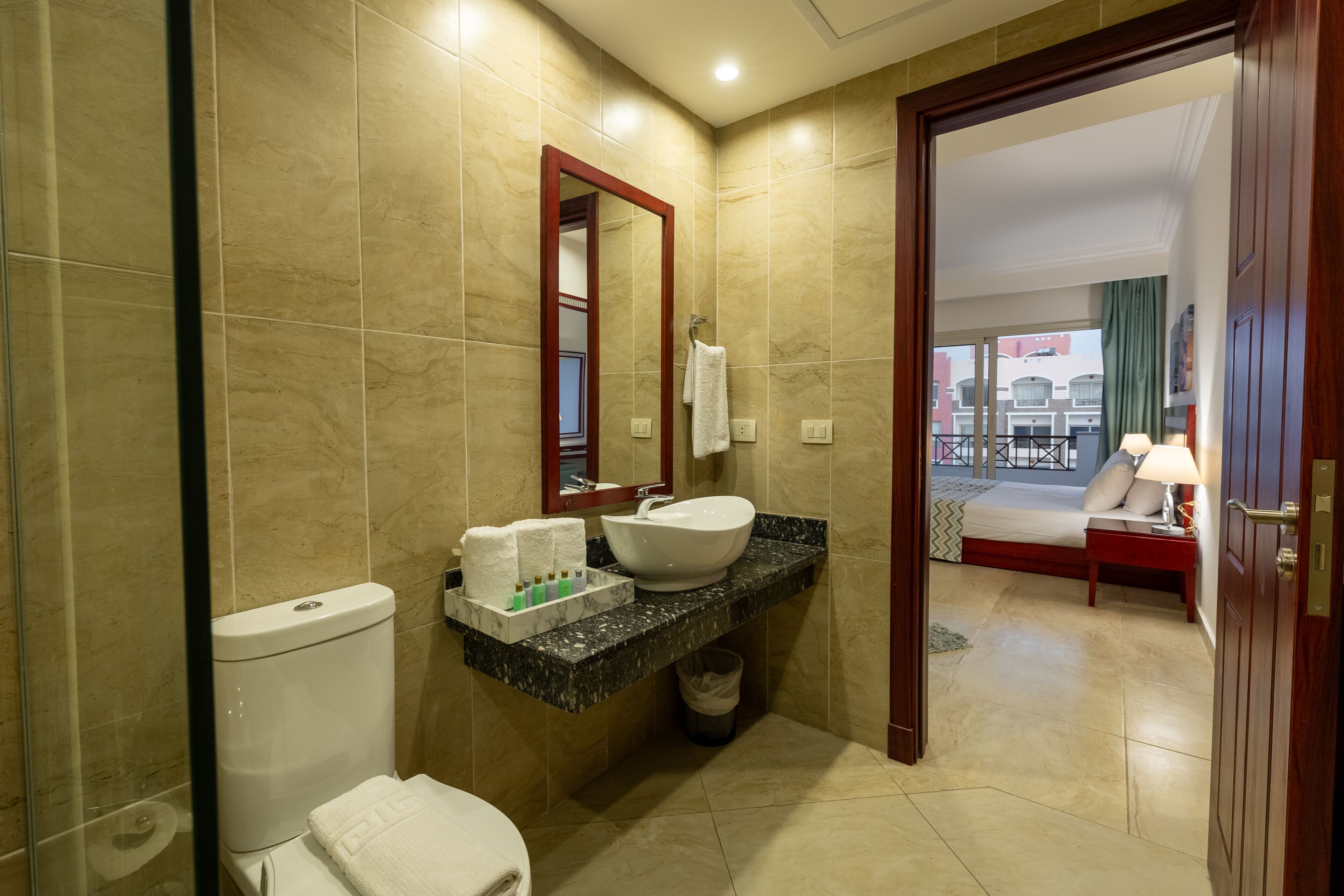 Casa de banho com sanita, lavatório e espelho. Há um porta-toalhas com toalhas e um contentor de lixo. O espaço tem uma porta que dá para outra área.