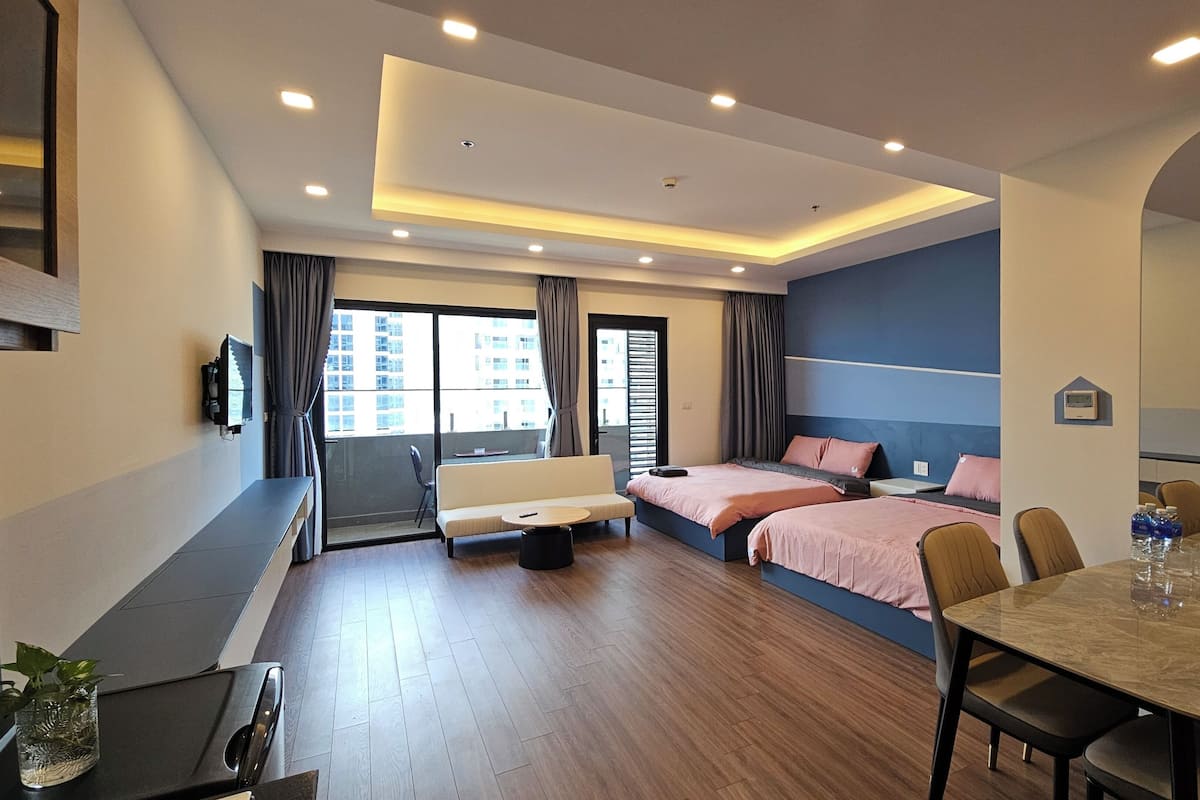 Quarto de hotel moderno com sofá, duas camas, mesa de jantar e varanda com cortinas.