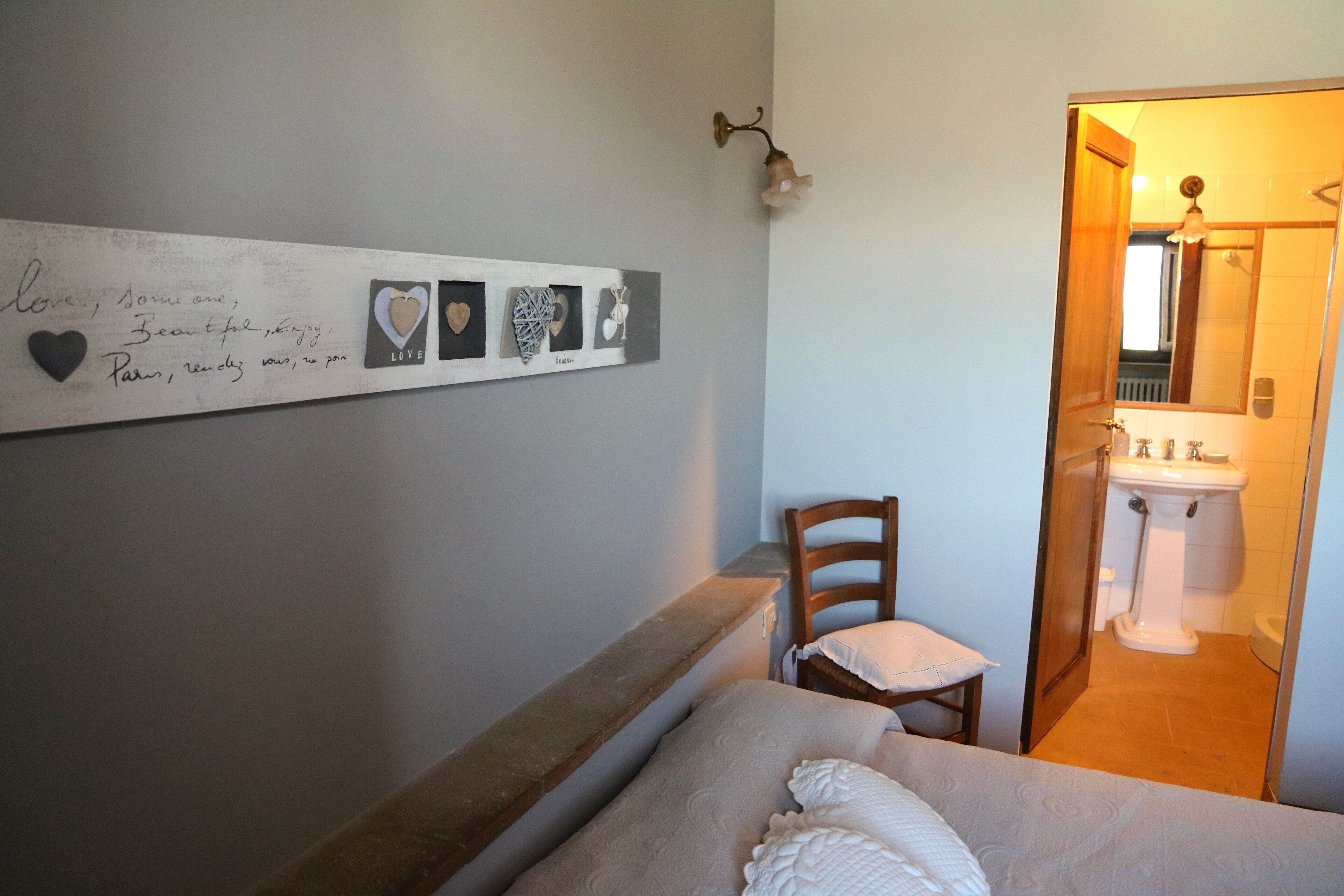 Quarto com cama, cadeira, mesa de cabeceira, prateleira na parede com objetos decorativos e banheiro visível através de uma porta aberta.