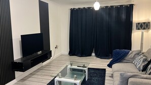 Uma sala de estar moderna com televisão de tela plana, mesa de centro de vidro, sofá e cortina escura.