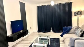 Uma sala de estar moderna com televisão de tela plana, mesa de centro de vidro, sofá e cortina escura.