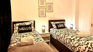 Quarto de hotel com duas camas, cada uma com colcha estampada e almofadas com texto. Há quadros nas paredes, um abajur e uma lâmpada pendurada no teto.