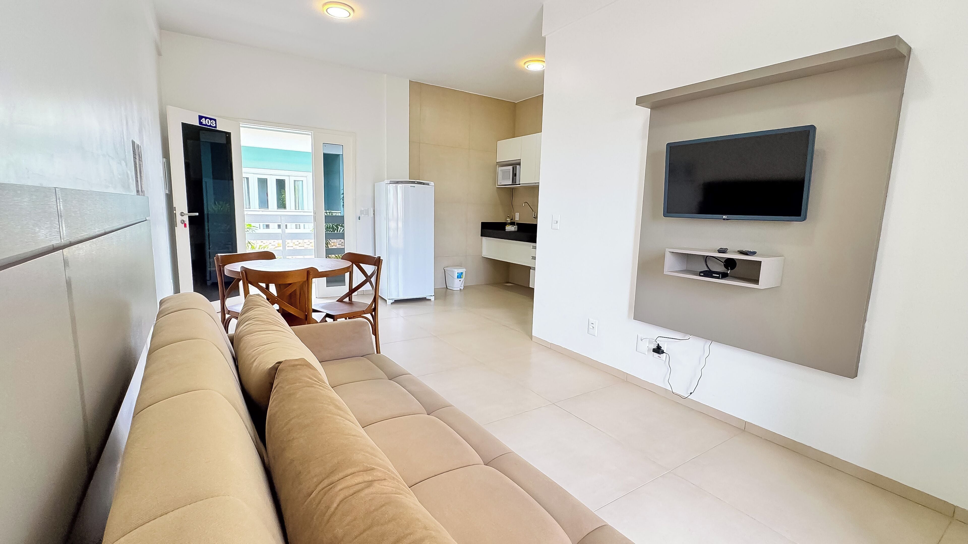 Uma sala moderna com um sofá bege, uma mesa de jantar com cadeiras, uma cozinha com micro-ondas e uma televisão na parede.