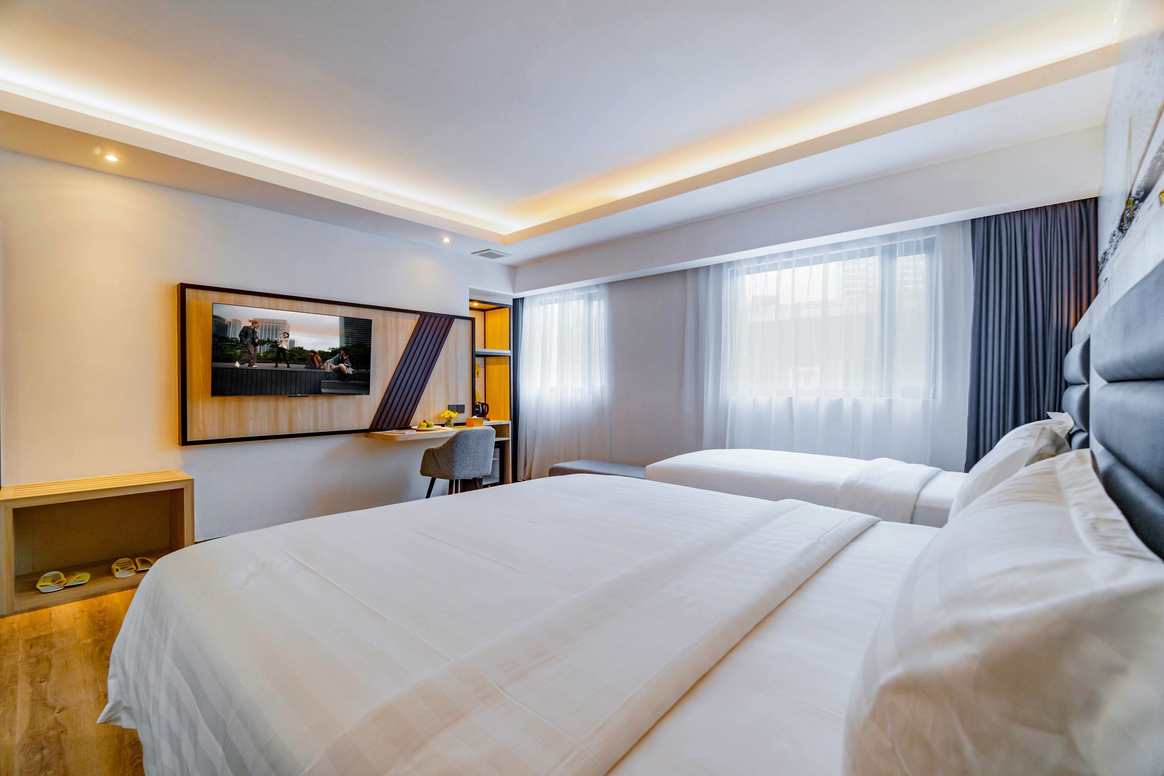 Quarto de hotel com duas camas, uma escrivaninha, uma cadeira, um pequeno armário com sapatos e uma janela grande com cortinas.