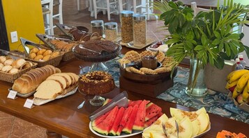 Mesa de buffet com diversos alimentos, incluindo pão, bolos e frutas.