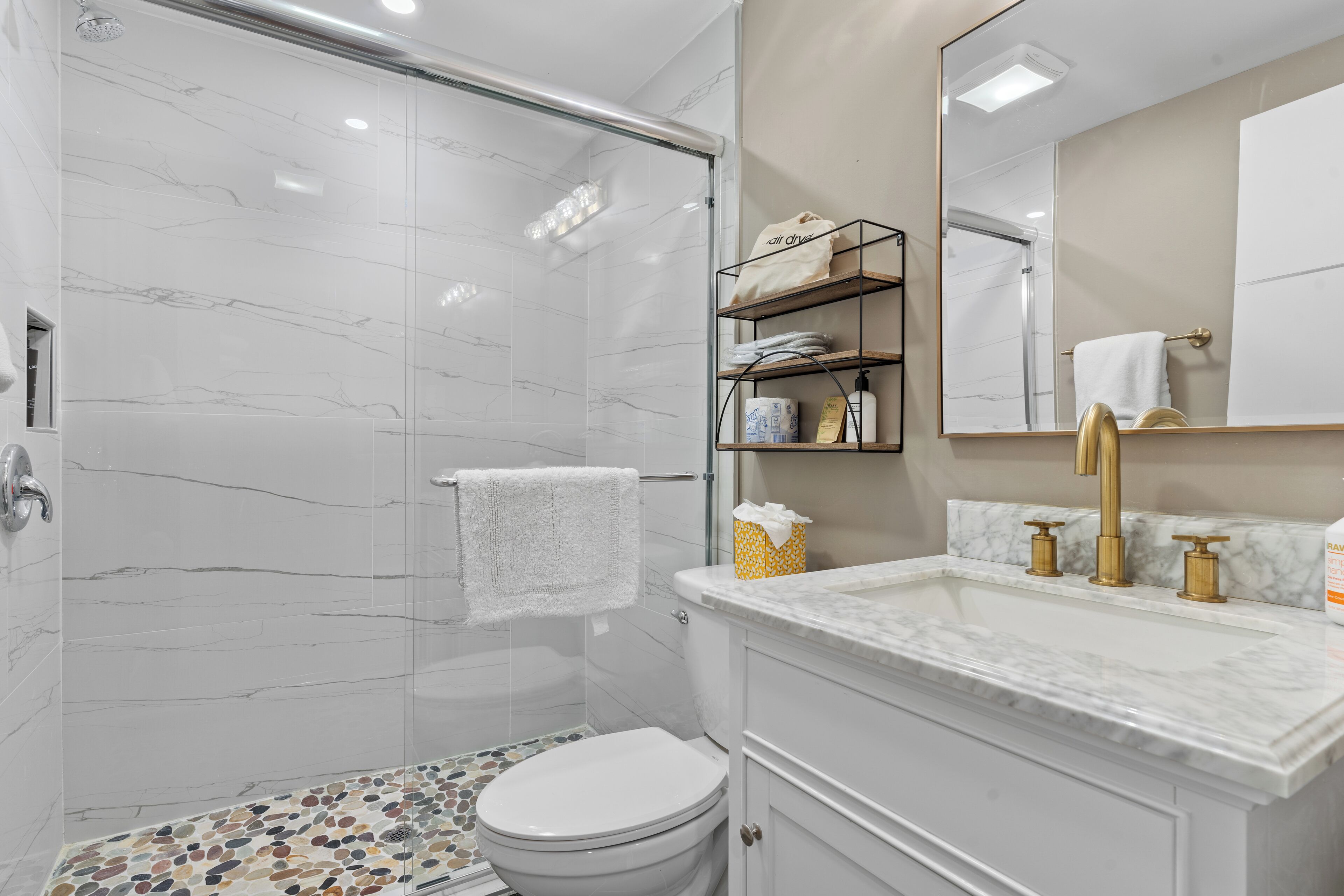 Uma casa de banho moderna com cabine de duche em vidro, chão em mosaico de mármore, sanita branca, toucador com torneira dourada e prateleira com produtos de higiene pessoal.