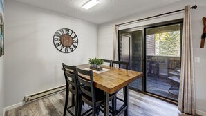 Uma área de refeições com mesa e cadeiras de madeira, um relógio na parede e uma porta de vidro deslizante que dá para um pátio externo.