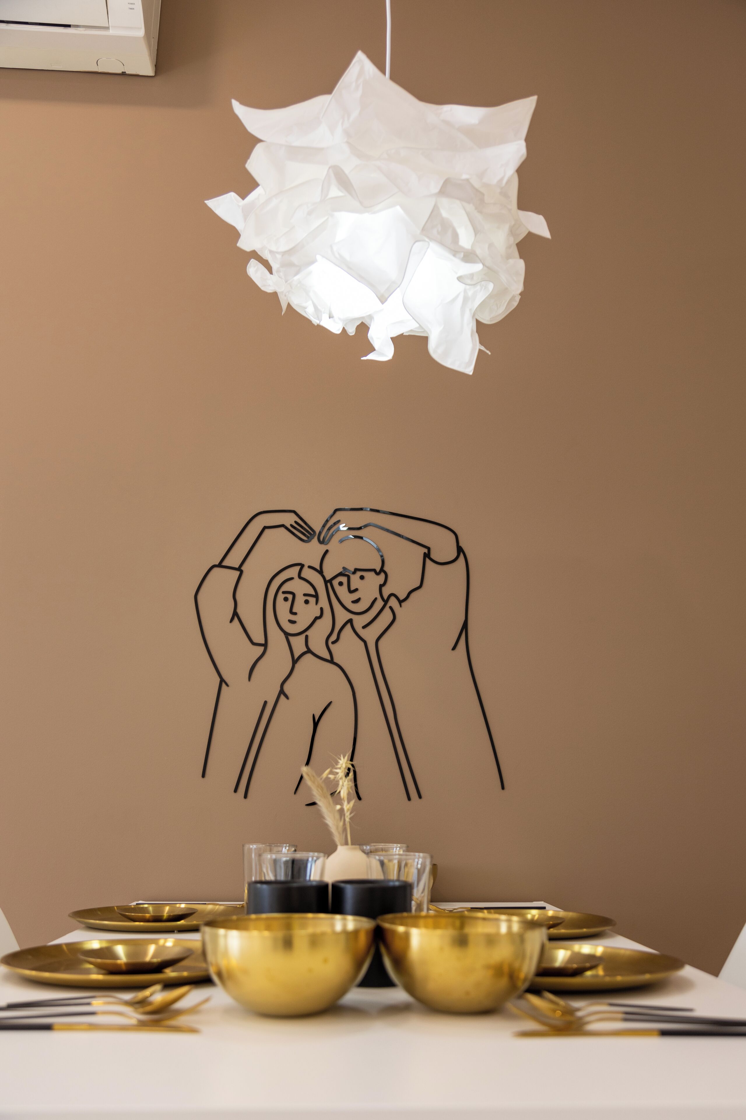 Uma mesa com tigelas douradas, um lanterna de papel branca e uma obra de arte na parede retratando duas pessoas formando um coração.
