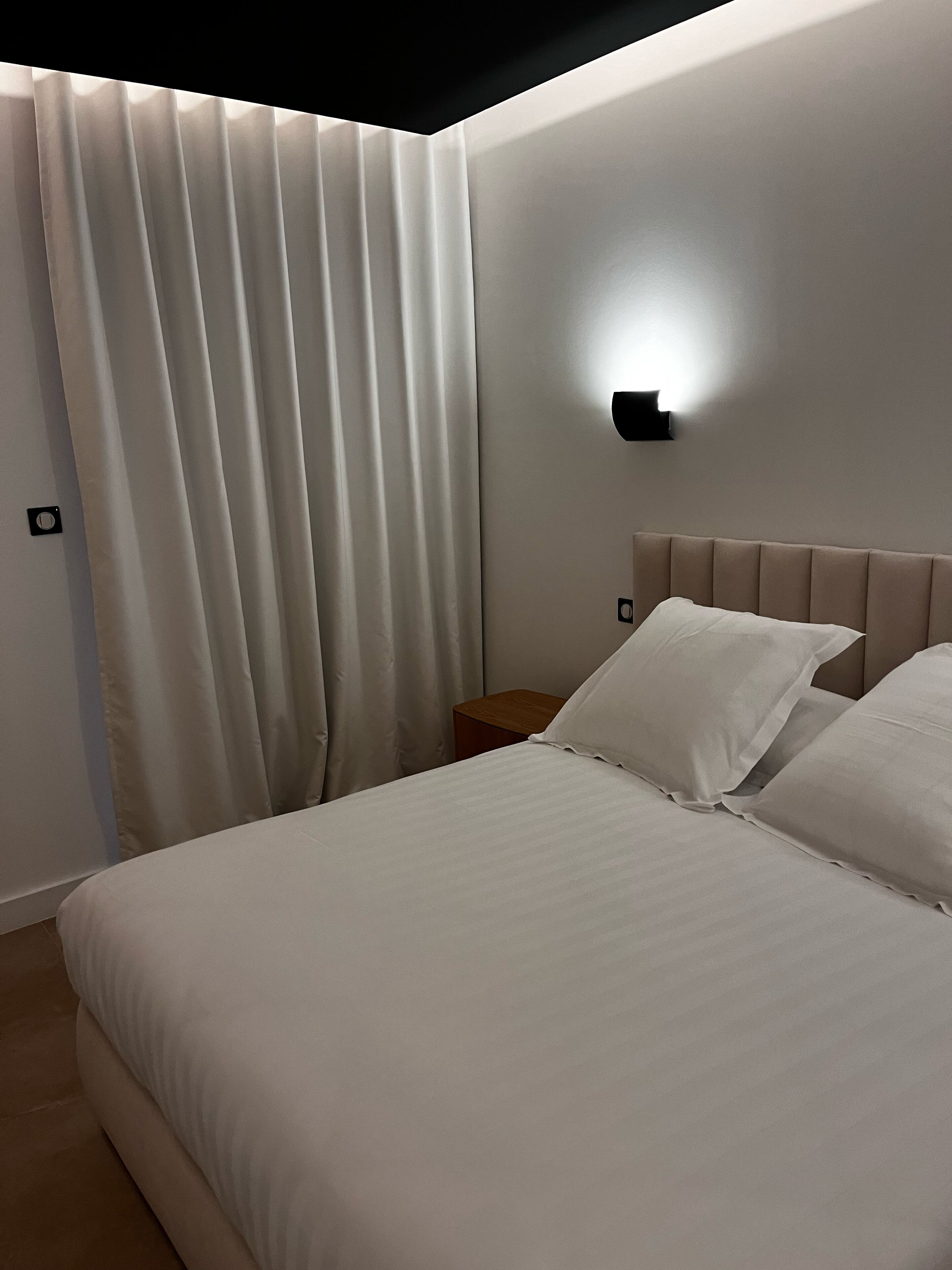 Uma cama bem arrumada com roupa de cama branca, cabeceira e mesa de cabeceira com abajur.
