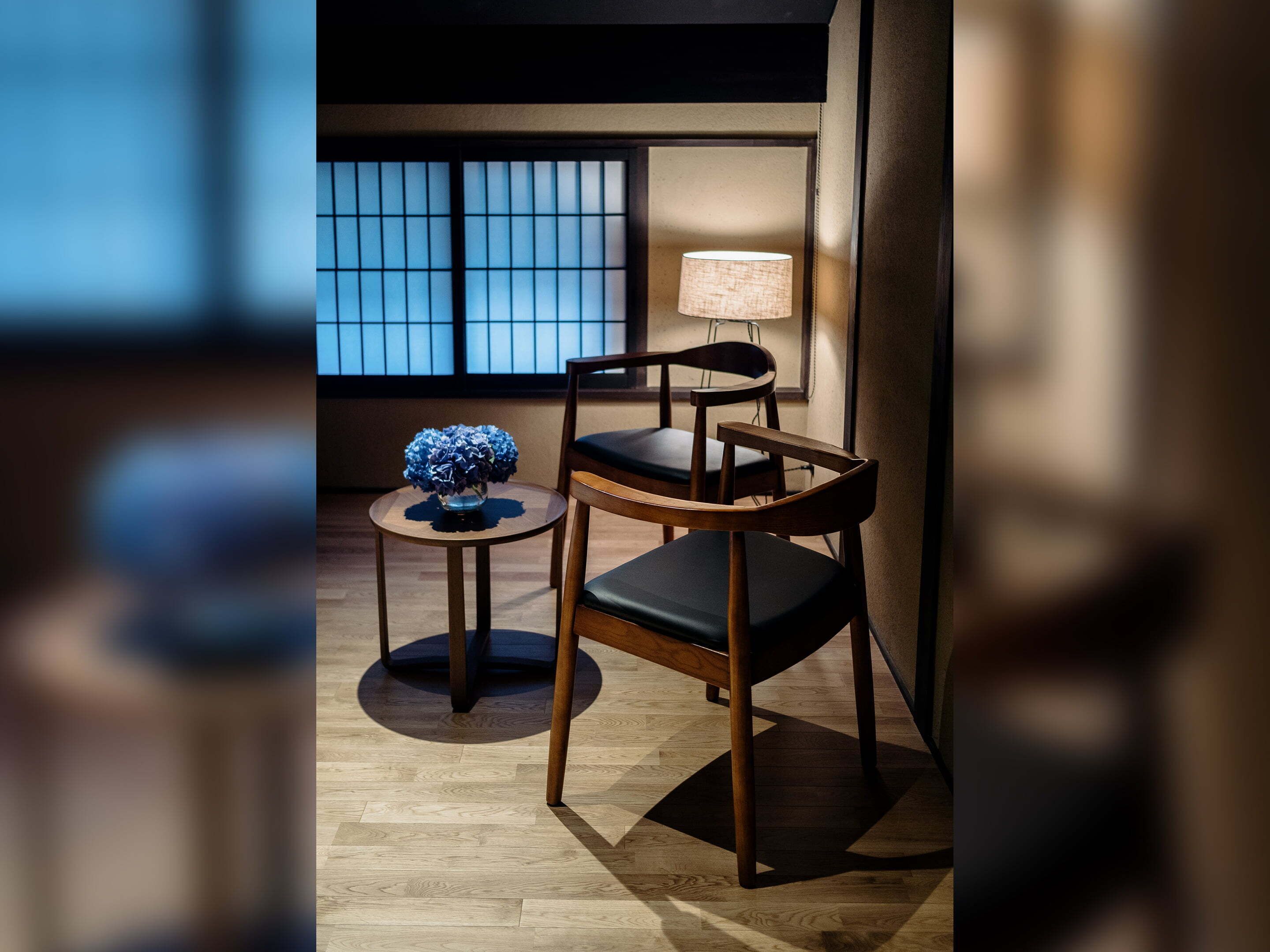 Quarto moderno com uma cadeira de madeira, uma mesa redonda pequena com um vaso de flores e uma luminária.
