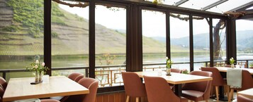 Um restaurante com vista para um rio e colinas. Mesas dispostas com cadeiras, e arranjos de flores sobre elas.