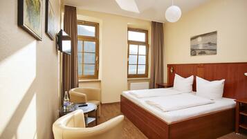 Quarto de hotel com cama, escrivaninha, cadeira e duas janelas.