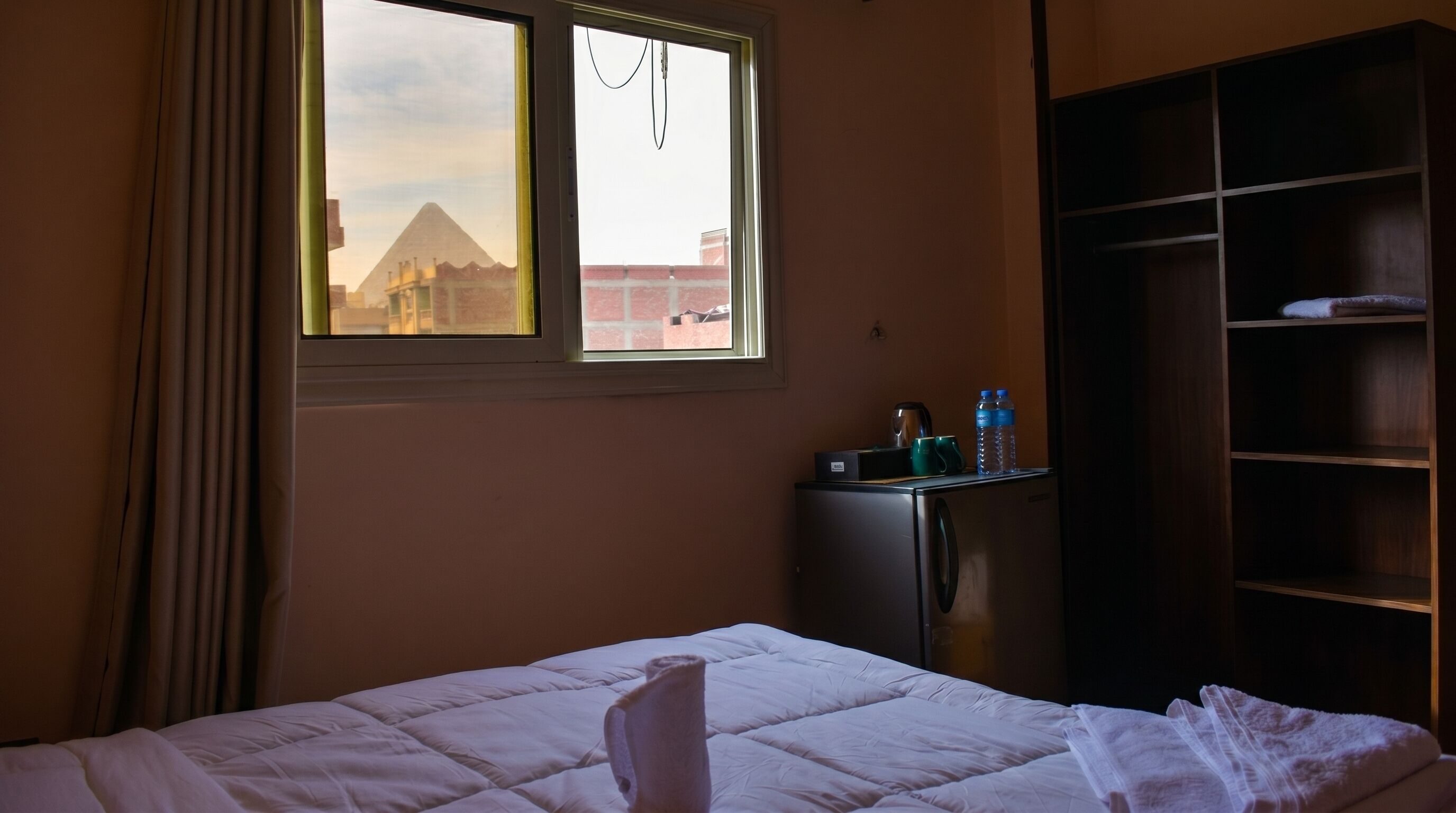 Quarto de hotel com cama, janela com vista para uma pirâmide, minibar e guarda-roupa.