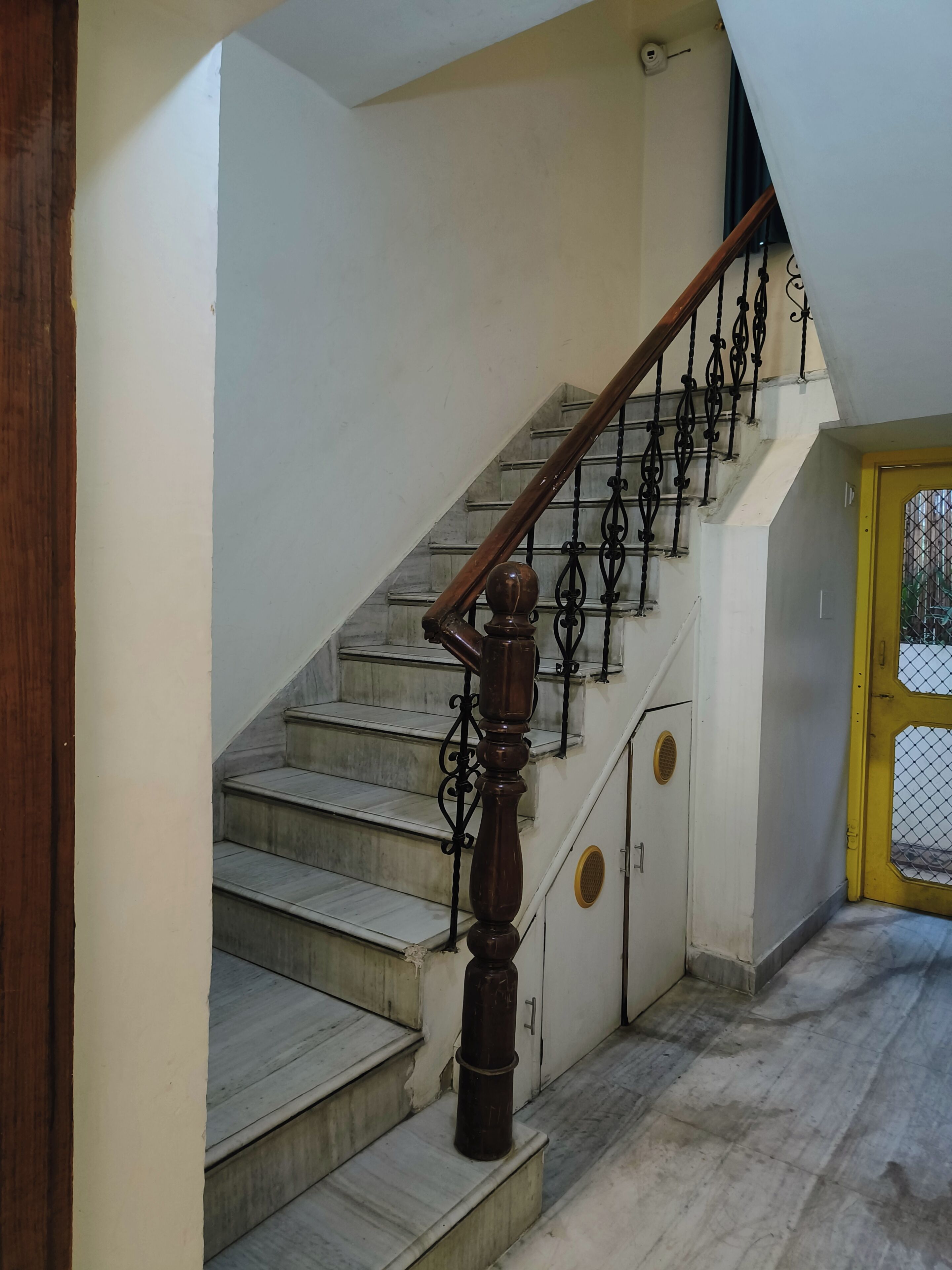 Uma escadaria com corrimão de madeira e balaústres pretos, conduzindo a um quarto com uma porta amarela e uma janela com grade de metal.