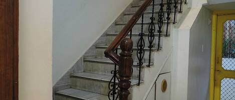 Uma escadaria com corrimão de madeira e balaústres pretos, conduzindo a um quarto com uma porta amarela e uma janela com grade de metal.