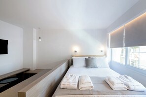 Quarto de hotel moderno com uma cama grande, mesas de cabeceira, televisão de tela plana e janela com persianas.