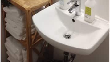 Uma casa de banho com uma pia branca, duas embalagens de sabonete líquido, um telefone preto, uma cesta com uma planta e uma prateleira de madeira com toalhas dobradas.