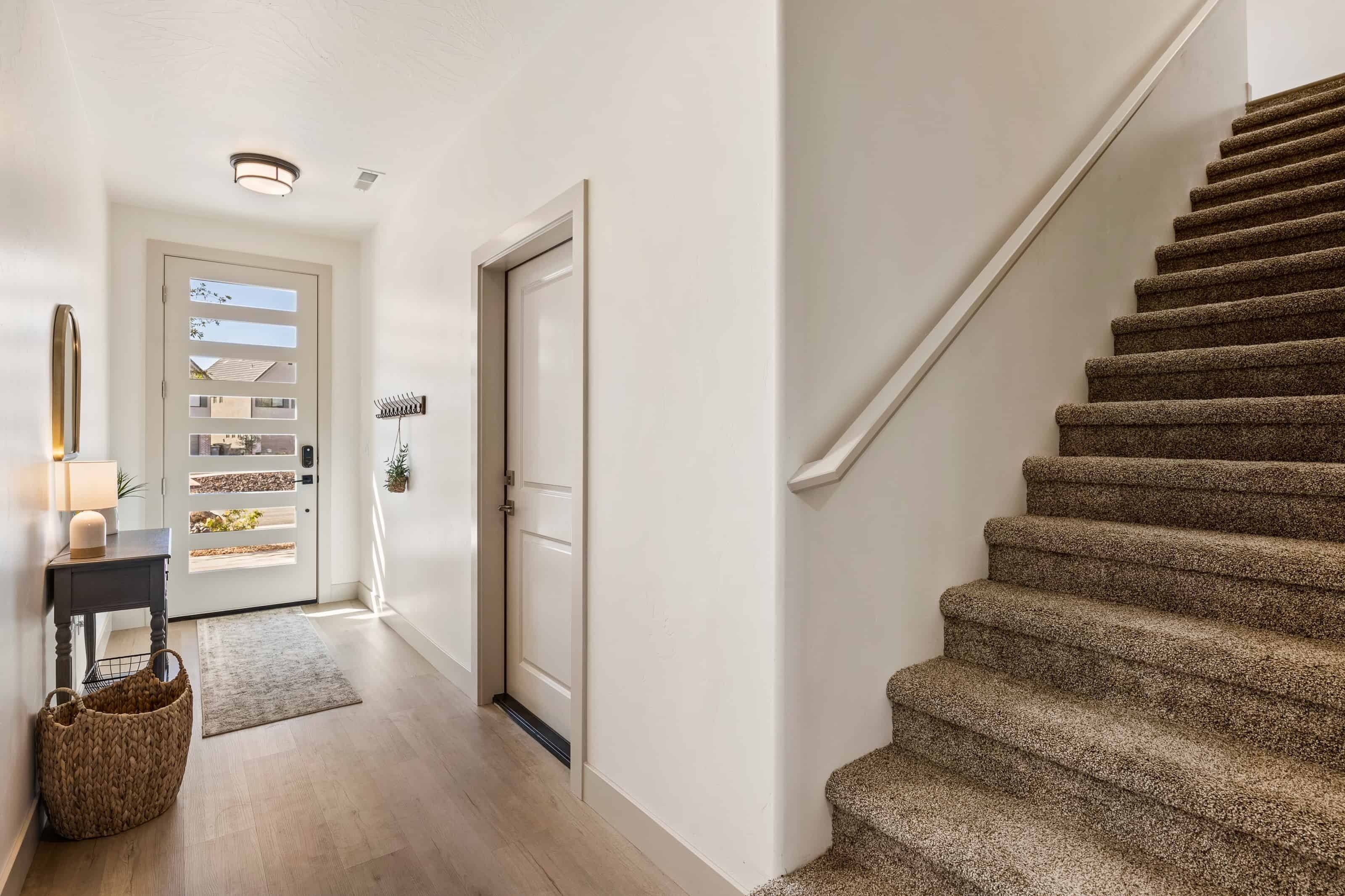 Um corredor com piso de madeira, escada com degraus carpetados, porta com painel de vidro e uma pequena mesa com luminária e cesto.