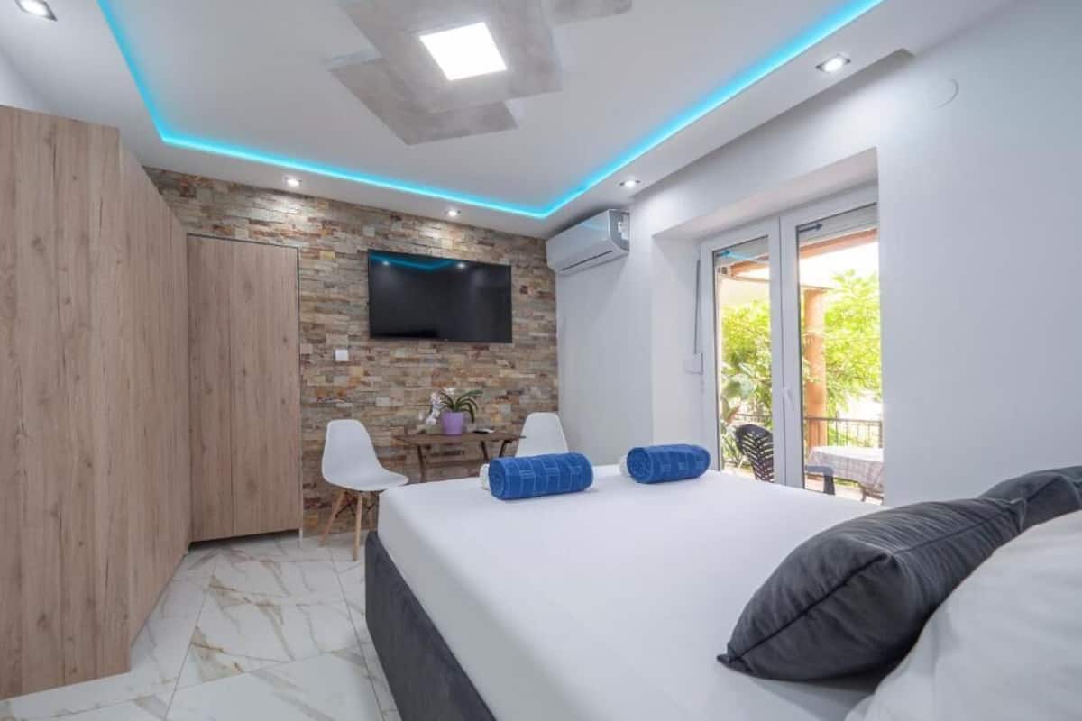 Um quarto moderno com uma cama grande, uma televisão presa a uma parede de pedra, uma escrivaninha com uma cadeira e uma porta de vidro deslizante para uma varanda.