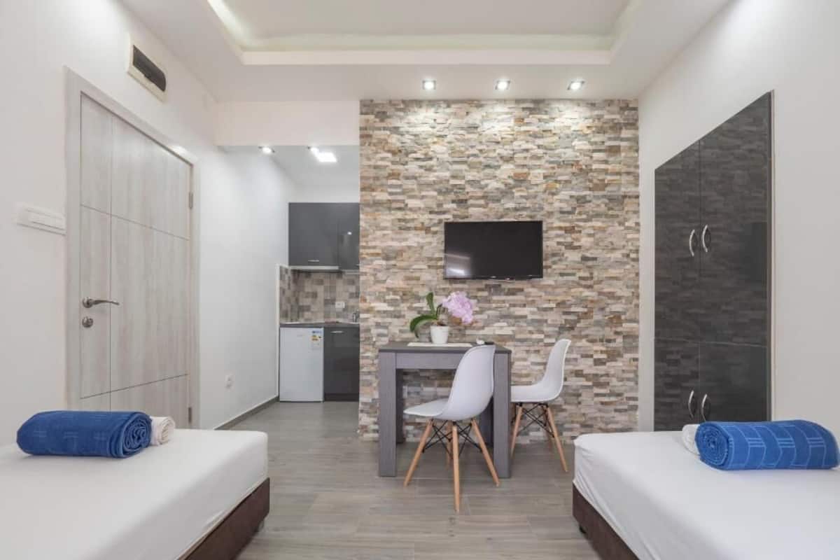 Quarto de hotel moderno com parede de destaque em pedra, TV de tela plana, escrivaninha com duas cadeiras e cama com toalhas enroladas.