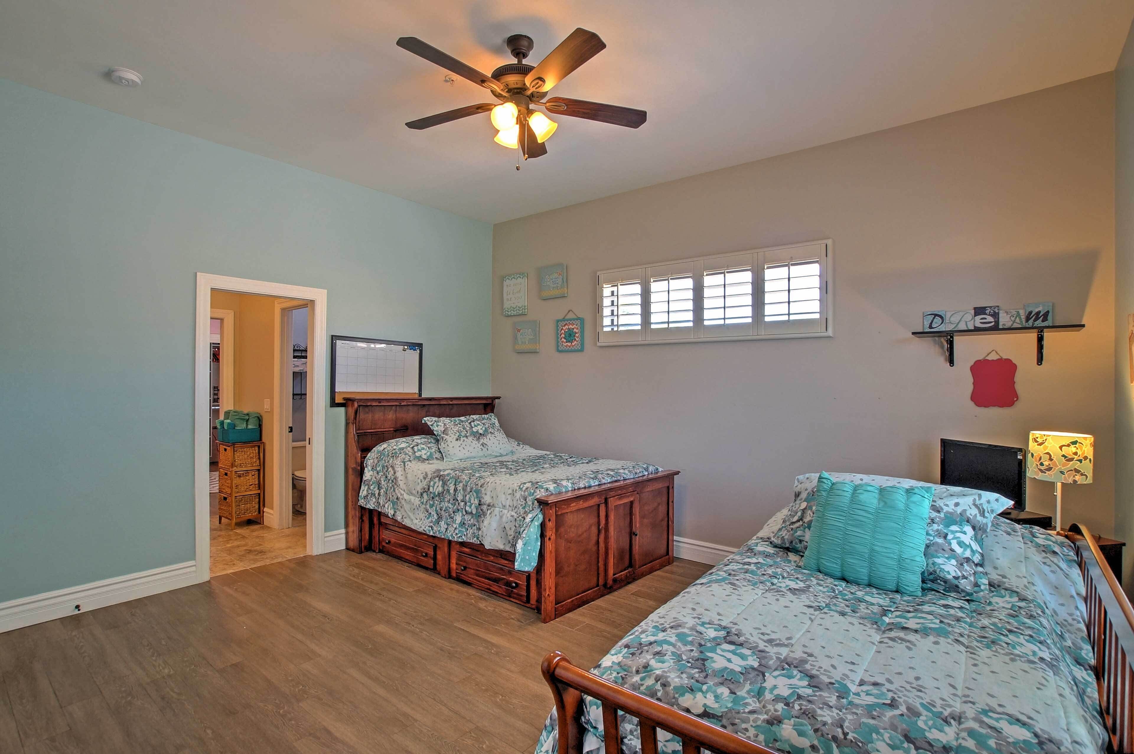 Quarto com cama de madeira, ventoinha no teto, janela com persianas e prateleira com objetos decorativos.
