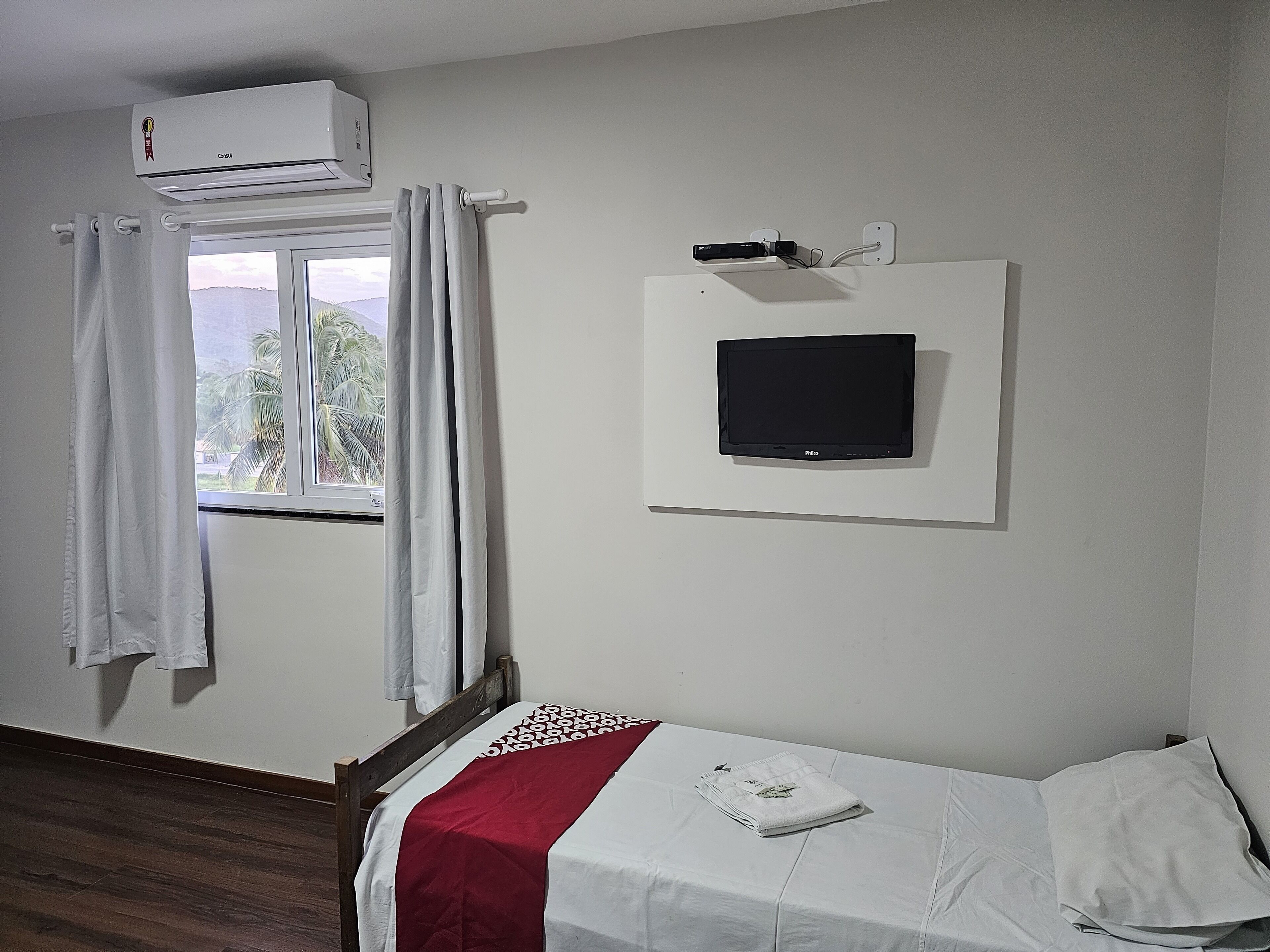 Quarto com cama, televisão na parede, ar condicionado e janela com cortinas.