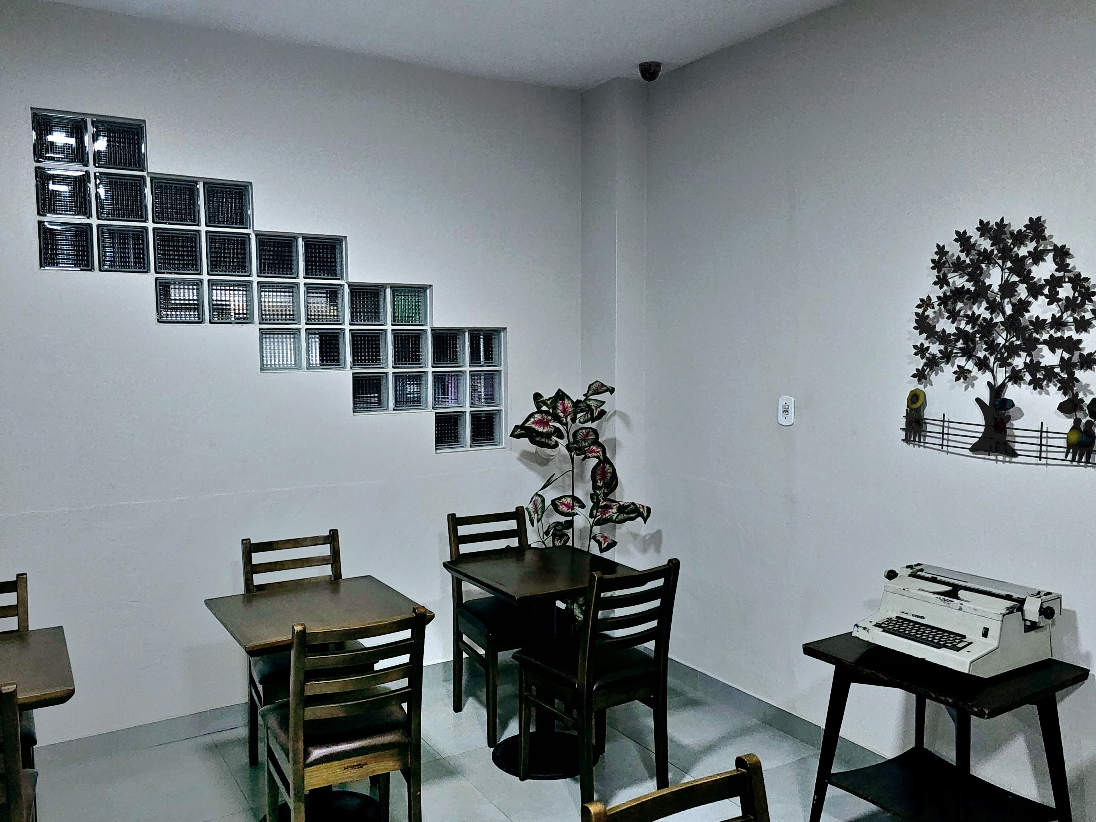 Um quarto com uma mesa de jantar, cadeiras, um vaso com planta, uma máquina de escrever e uma parede com padrão geométrico e um enfeite de árvore.