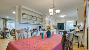 Uma sala de jantar com uma mesa posta para quatro pessoas, rodeada por cadeiras de madeira. Um lustre pendurado acima da mesa, e um vaso com flores sobre ela.