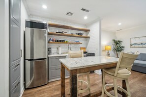 Uma cozinha moderna com geladeira de aço inoxidável, prateleiras de madeira, bancada de mármore e um espaço de jantar com mesa e cadeiras de madeira.