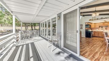 Uma área externa coberta com um deck de madeira, um espaço de estar com cadeiras e vista para uma cozinha com armários de madeira e um lustre.