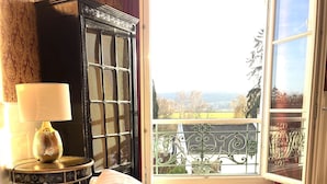 Quarto com cama, mesa de cabeceira com abajur e vista para uma varanda com guarda-corpo.