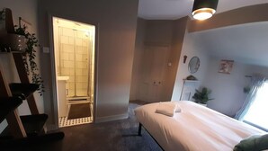 Quarto com cama, porta para um banheiro, escrivaninha pequena e prateleira com plantas.