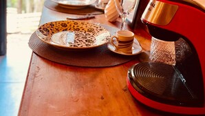 Uma mesa de madeira com uma cafeteira, um copo de água, um vaso com flores e um quadro emoldurado.