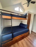 Uma cama beliche com estrutura de madeira e gavetas, um ventilador de teto e uma janela com vista.
