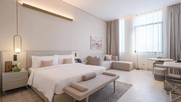 Quarto de hotel moderno com uma cama grande, área de estar, escrivaninha e vista para o exterior através de amplas janelas.