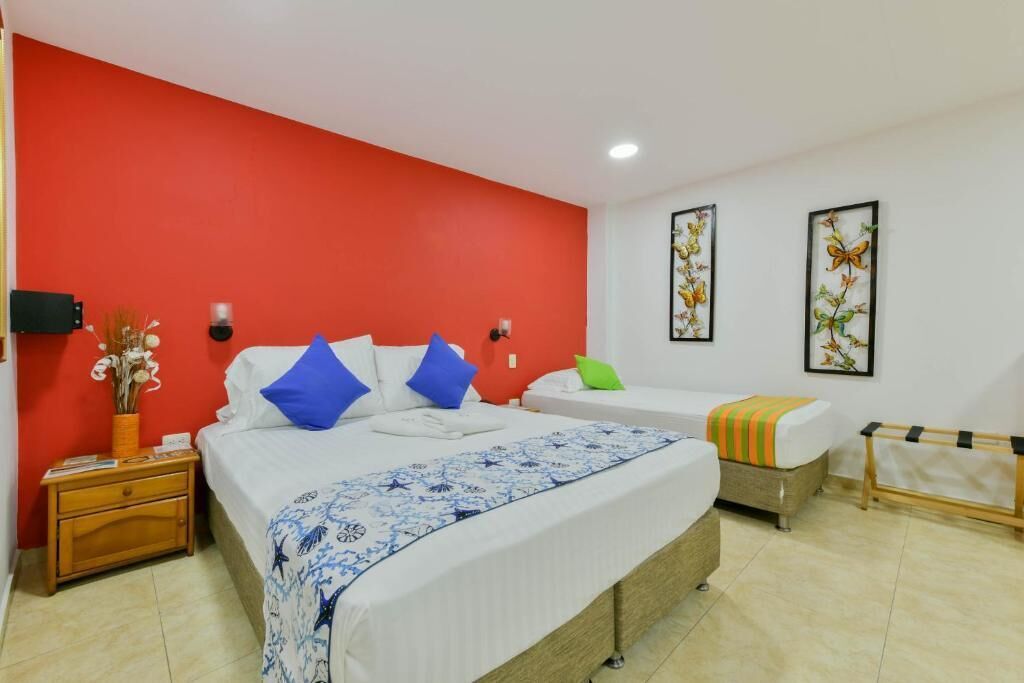Quarto de hotel com uma parede de destaque vermelha, uma cama grande com roupa de cama azul e branca, duas camas menores, uma mesa de cabeceira de madeira com um vaso de flores e dois quadros florais emoldurados na parede.