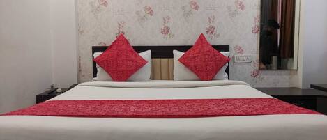 Quarto de hotel com cama, duas almofadas vermelhas, colcha branca e papel de parede floral.